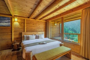 Quarto de cabana de madeira com cama, mesa de cabeceira, banco e amplas janelas com vista para a floresta.