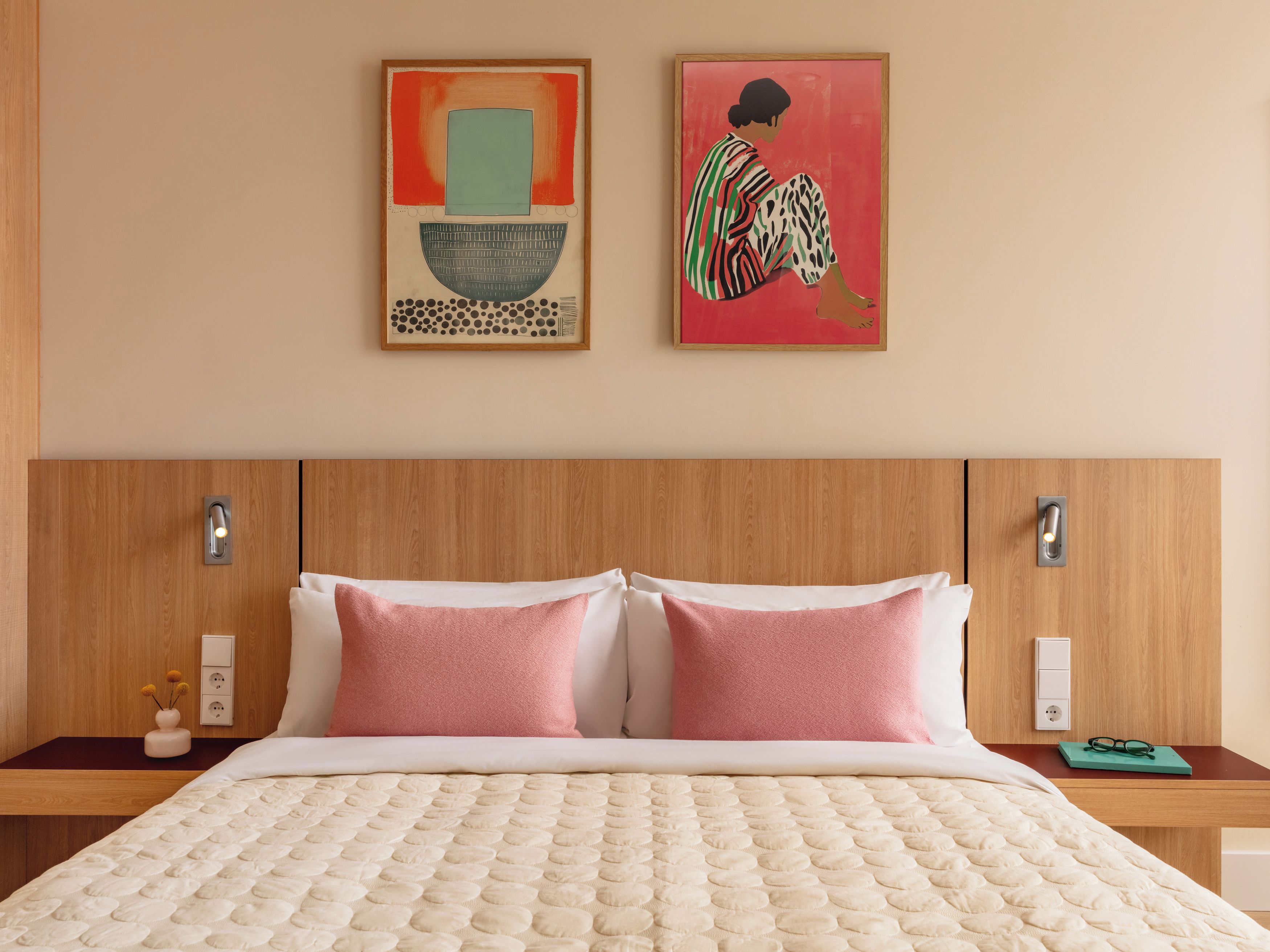 Quarto de hotel com cabeceira de madeira, cama com cobertura acolchoada, duas almofadas cor-de-rosa e dois interruptores de luz fixados na parede.