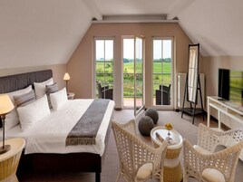 Quarto de hotel com cama, escrivaninha, televisão, cadeiras de vime e varanda com vista para o campo.