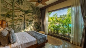 Quarto com cama grande, ventilador de teto e vista para um jardim tropical pela janela.