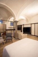 Quarto de hotel moderno com uma cama grande, área de estar com sofá e cadeiras, televisão e cozinha.