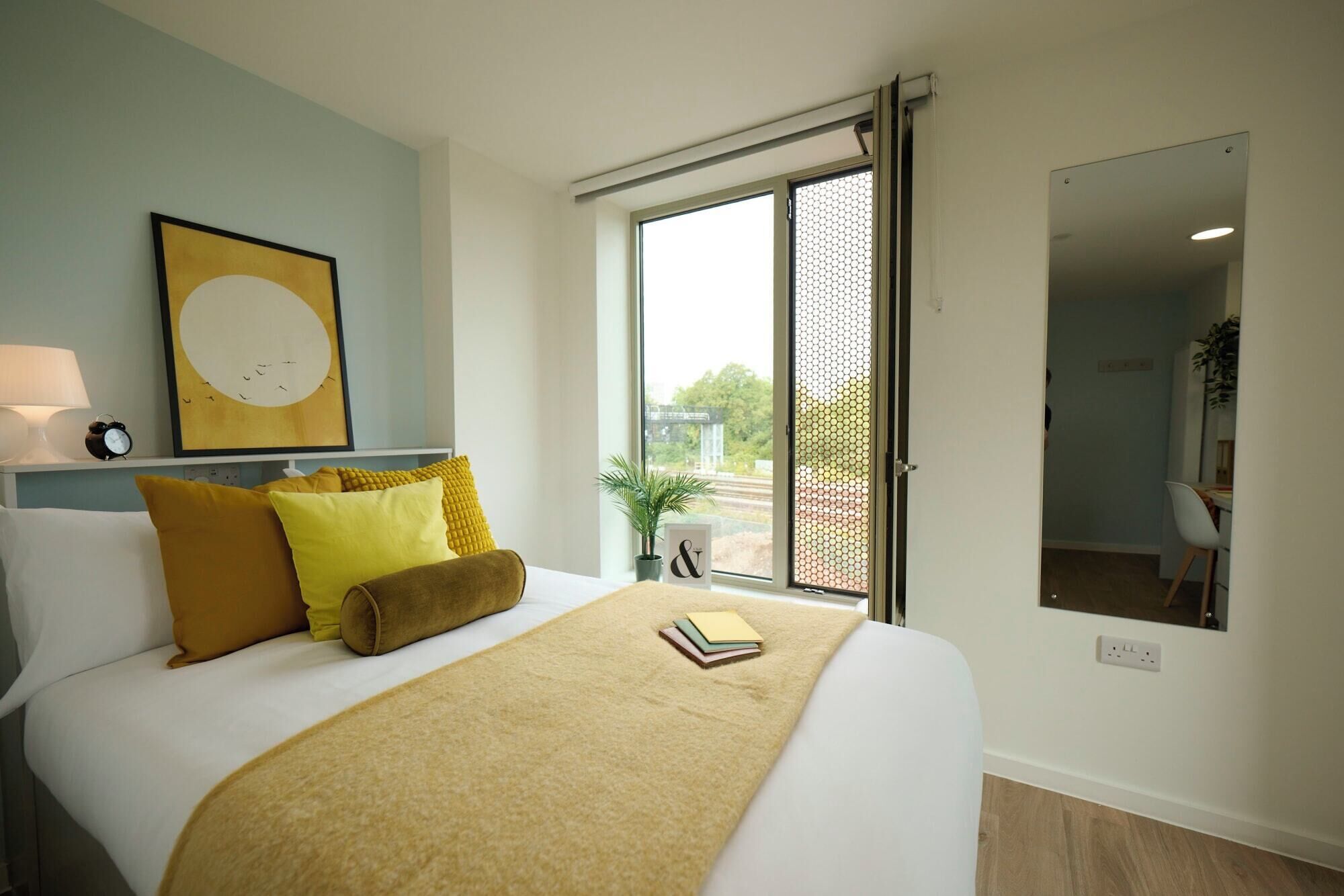 Quarto moderno com cama grande, mesa de cabeceira com luminária, quadro na parede e porta de vidro deslizante para uma varanda.