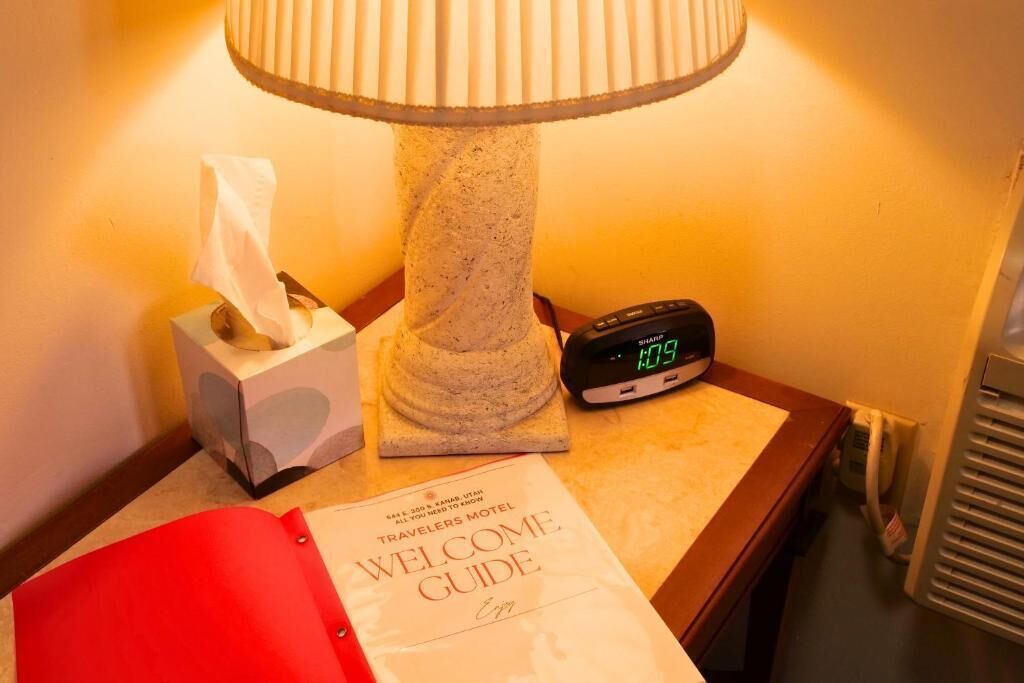 Um guia de boas-vindas, um lençoeiro, um relógio digital e uma luminária em uma mesa de cabeceira.
