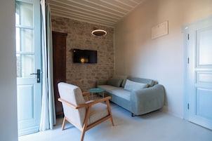 Uma sala de estar com parede de pedra, uma cadeira de madeira, um sofá claro, uma mesinha redonda e uma televisão fixada na parede.