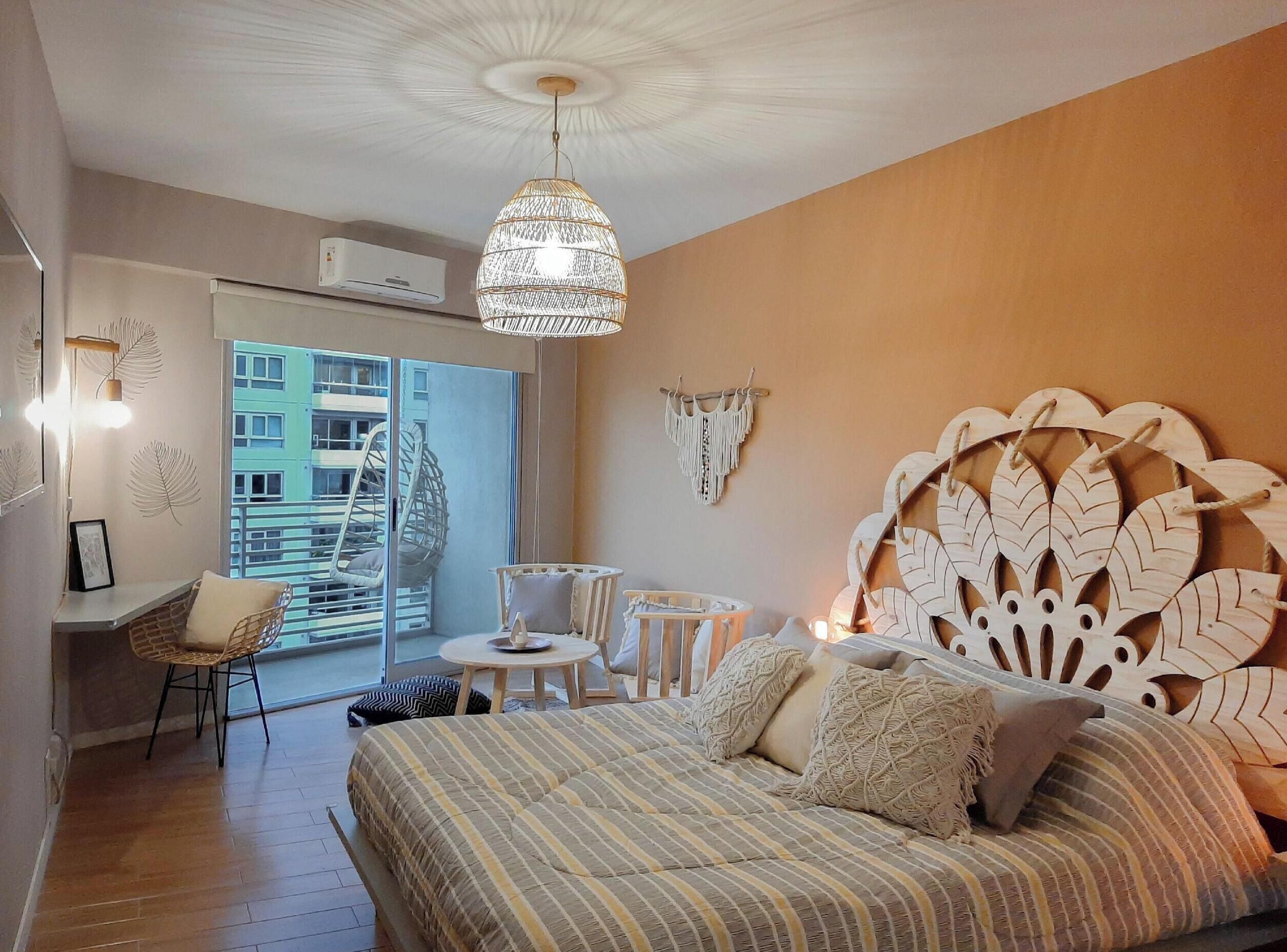 Quarto com cama, luminária pendente, uma cadeira, mesa pequena e um objeto decorativo na parede.