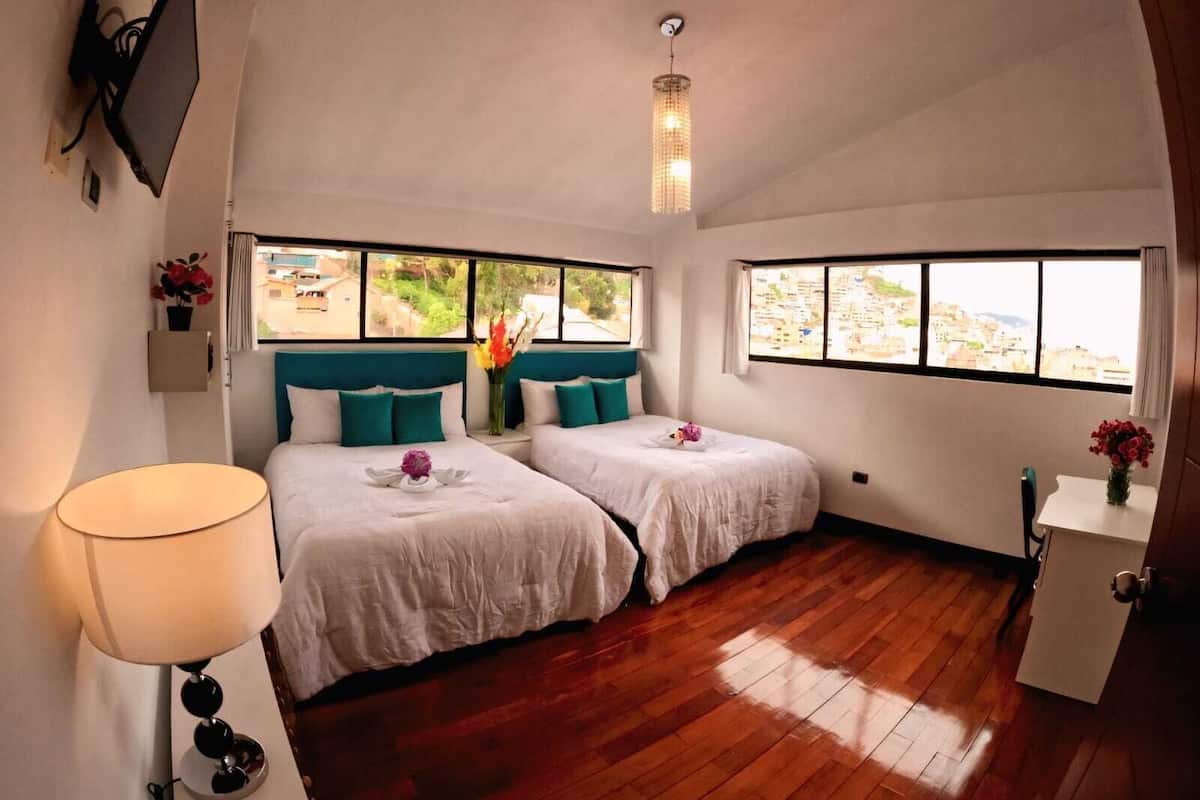 Quarto com duas camas, uma escrivaninha, uma luminária e vista para a cidade.