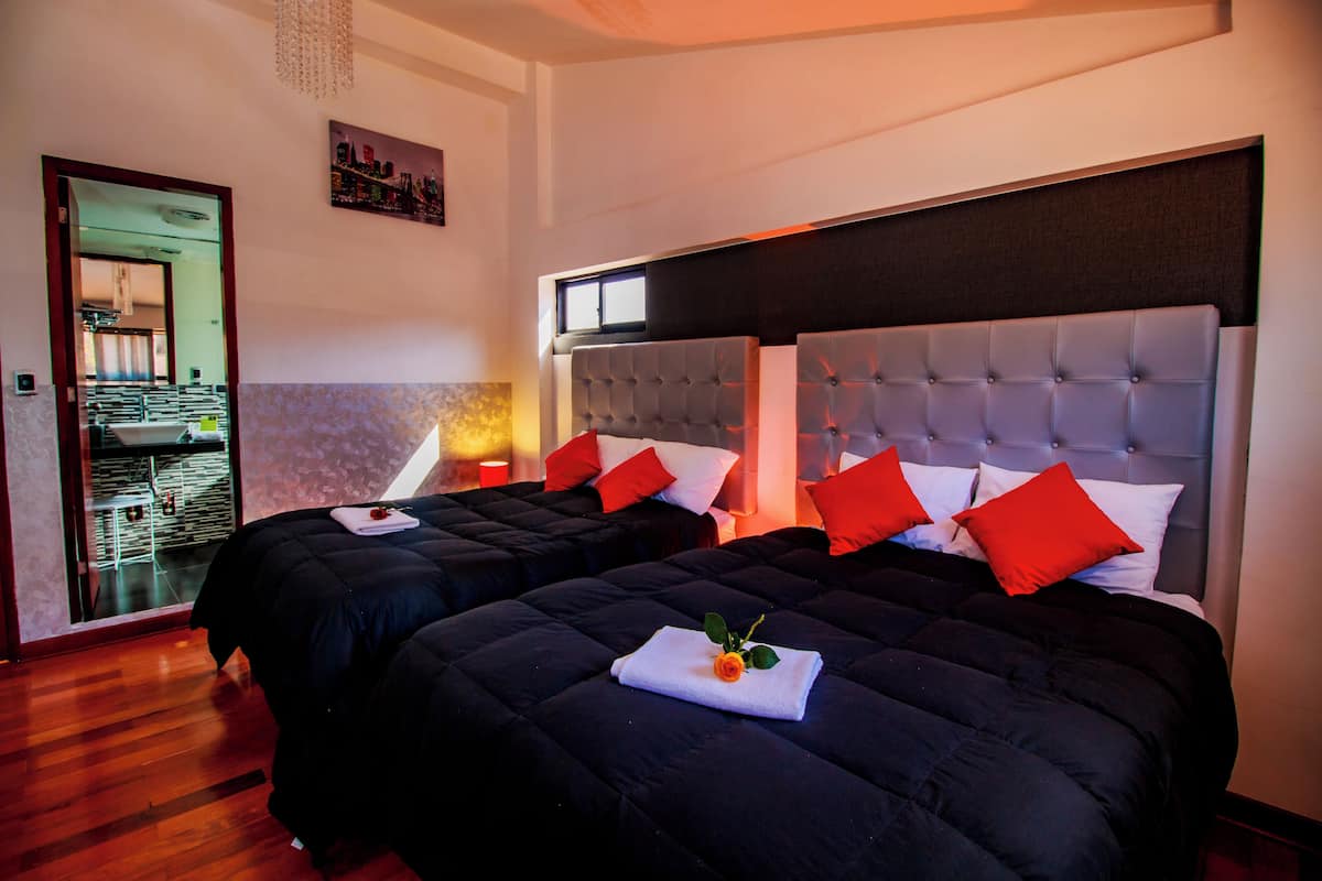 Quarto de hotel com duas camas, cada uma com cabeceira estofada, almofadas brancas e vermelhas e uma pequena flor laranja sobre uma toalha dobrada.