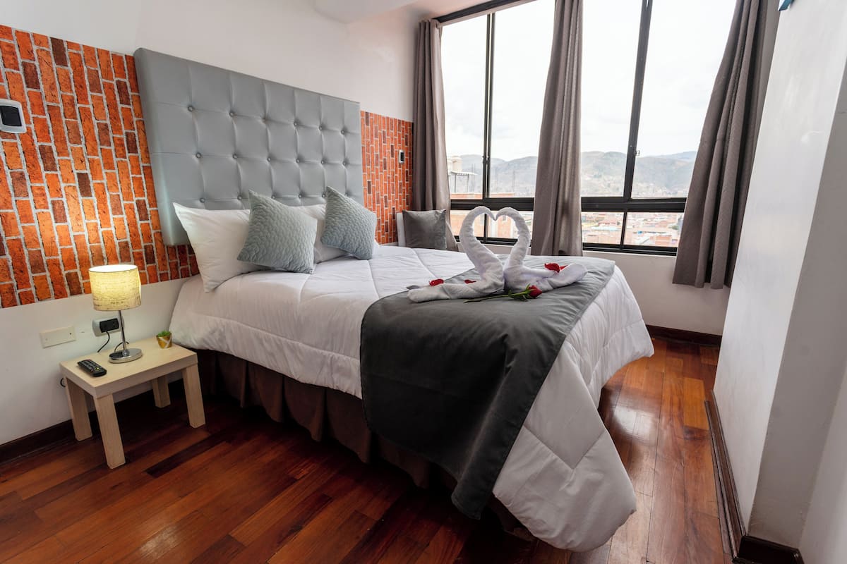 Quarto de hotel com parede em padrão tijolo, cabeceira estofada, cama com roupa de cama branca, mesa de cabeceira com abajur e vista da cidade através de grandes janelas.