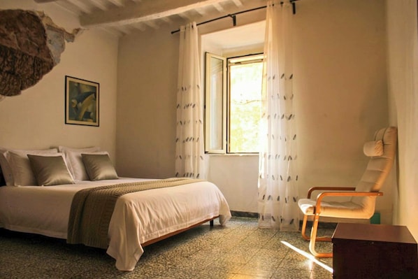 Um quarto com uma cama, uma cadeira, uma janela com cortinas e um quadro na parede.