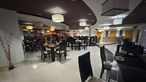 Uma ampla área de refeições com várias mesas e cadeiras, um espaço de bar central e uma cozinha visível ao fundo.