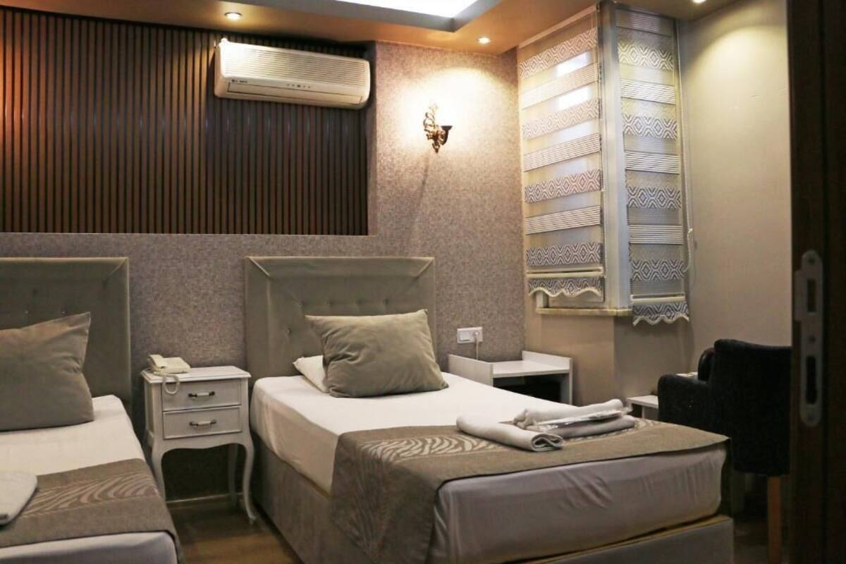 Quarto de hotel com duas camas, mesa de cabeceira com abajur, ar-condicionado e janela com persianas estampadas.