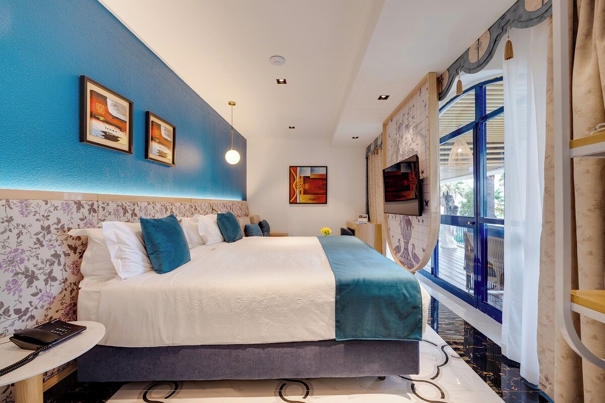 Quarto de hotel moderno com uma cama grande, papel de parede floral, uma escrivaninha com telefone e uma janela com cortinas.