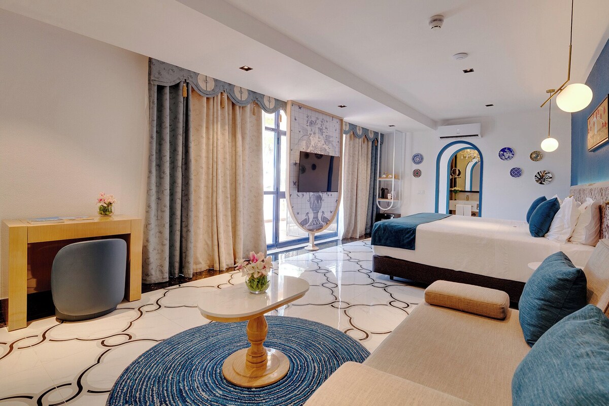 Quarto de hotel com cama, sofá, mesinha, cadeira e escrivaninha. Tem uma janela com cortinas, um espelho e uma parede decorada com pratos.