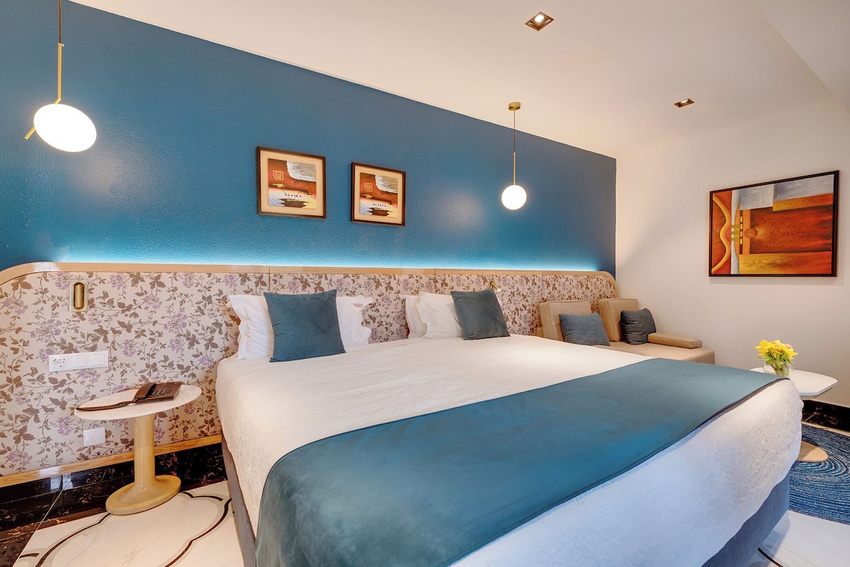 Quarto de hotel com uma cama grande, uma mesa lateral redonda, um telefone e um quadro na parede.