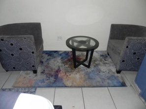 Um quarto pequeno com dois sofás cinzas e almofadas estampadas, uma mesa de vidro redonda e um tapete colorido.