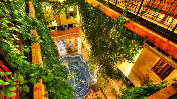 Um pátio interno com uma fonte em azulejos, plantas em vasos e um corrimão de madeira.