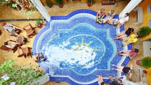 Uma piscina circular com design em mosaico, cercada por áreas de estar e vasos com plantas.