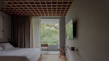 Quarto com cama, cadeira, mesa pequena, TV e varanda com vista para área verde.