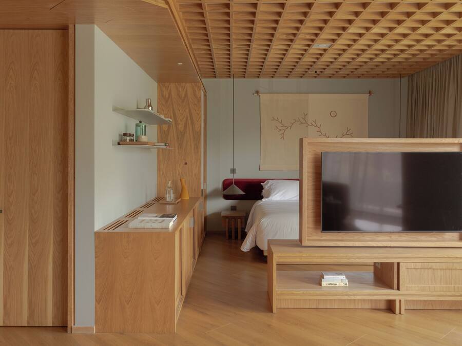 Quarto de hotel moderno com teto de madeira, cama com roupa de cama branca, televisão em suporte de madeira e uma pequena escrivaninha de madeira com cadeira.