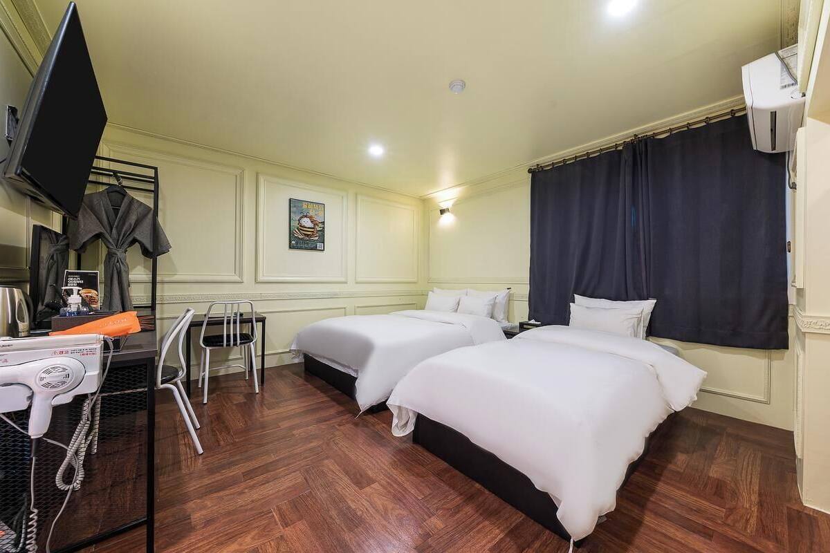 Quarto de hotel com duas camas, uma mesa com telefone e secador de cabelo, uma cadeira e uma janela grande com cortinas.