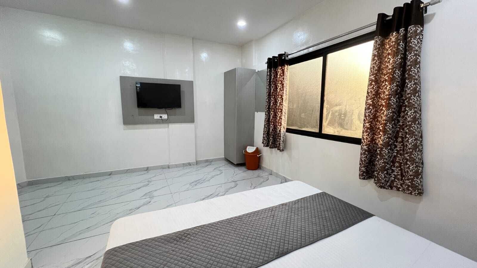 Quarto com cama, televisão fixada na parede, janela com cortinas e piso cerâmico.