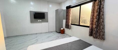 Quarto com cama, televisão fixada na parede, janela com cortinas e piso cerâmico.