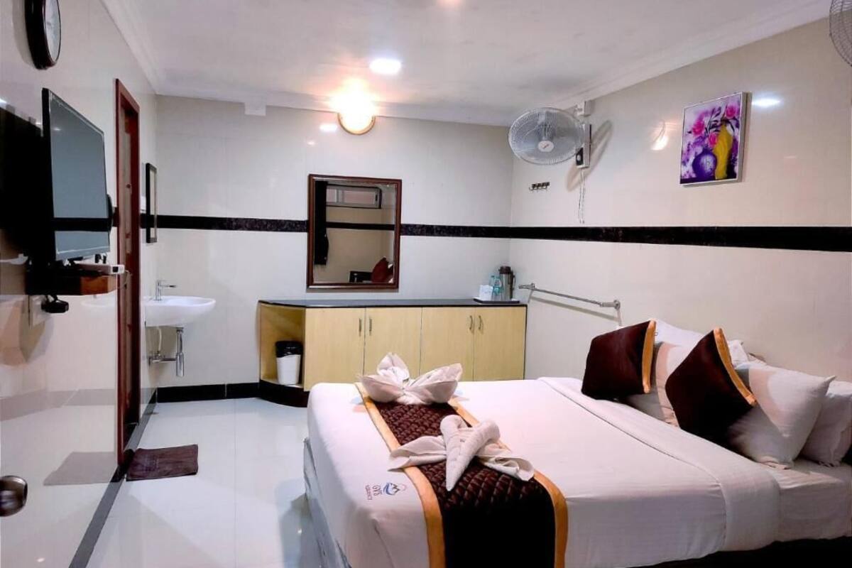 Quarto de hotel com cama, mesa de cabeceira, espelho, pia, armário, ventilador e relógio.