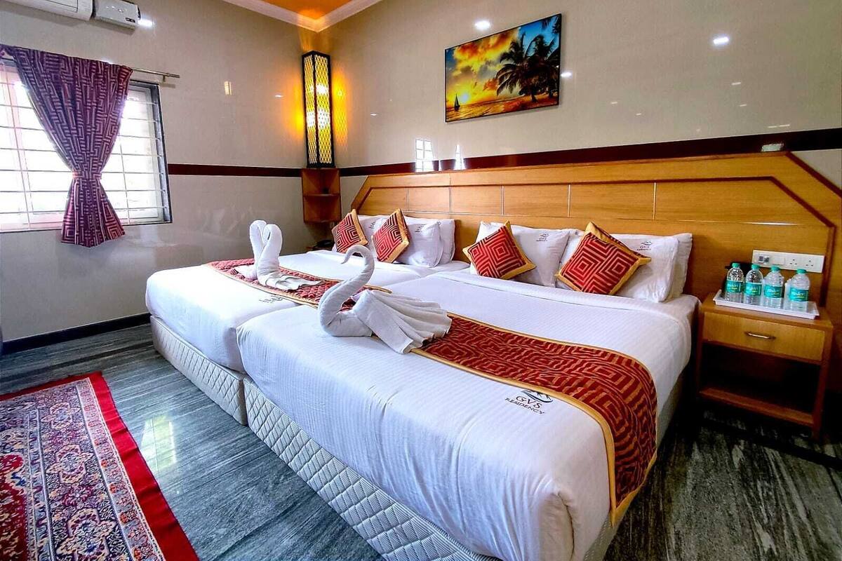 Quarto de hotel com duas camas, uma janela grande com cortinas, um quadro na parede e uma mesa de cabeceira com garrafas de água.