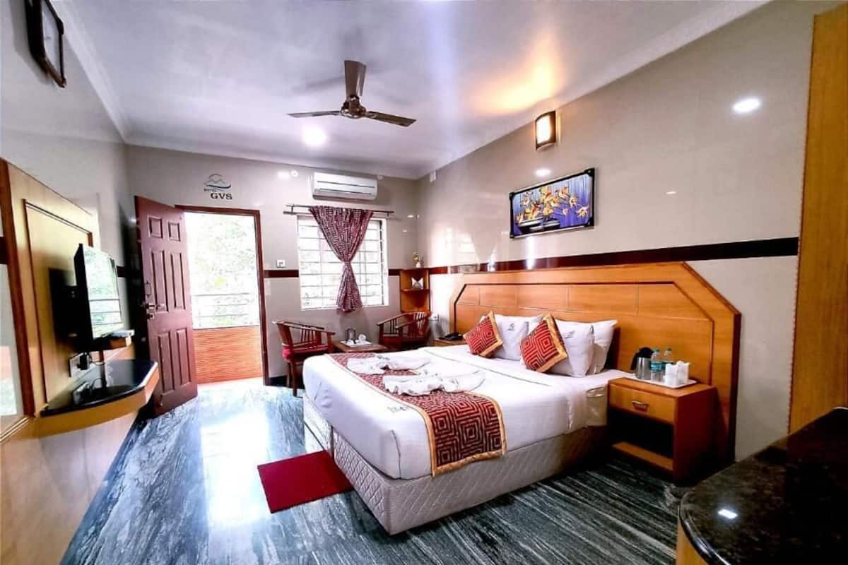 Quarto de hotel com uma cama grande, cabeceira de madeira, mesa de cabeceira, televisão, ventoinha no teto e janela com cortinas.
