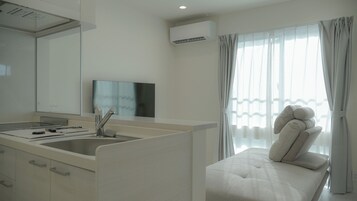Quarto de hotel moderno com cozinha compacta, espelho grande, televisão de tela plana, sofá com almofadas e janela com cortinas translúcidas.