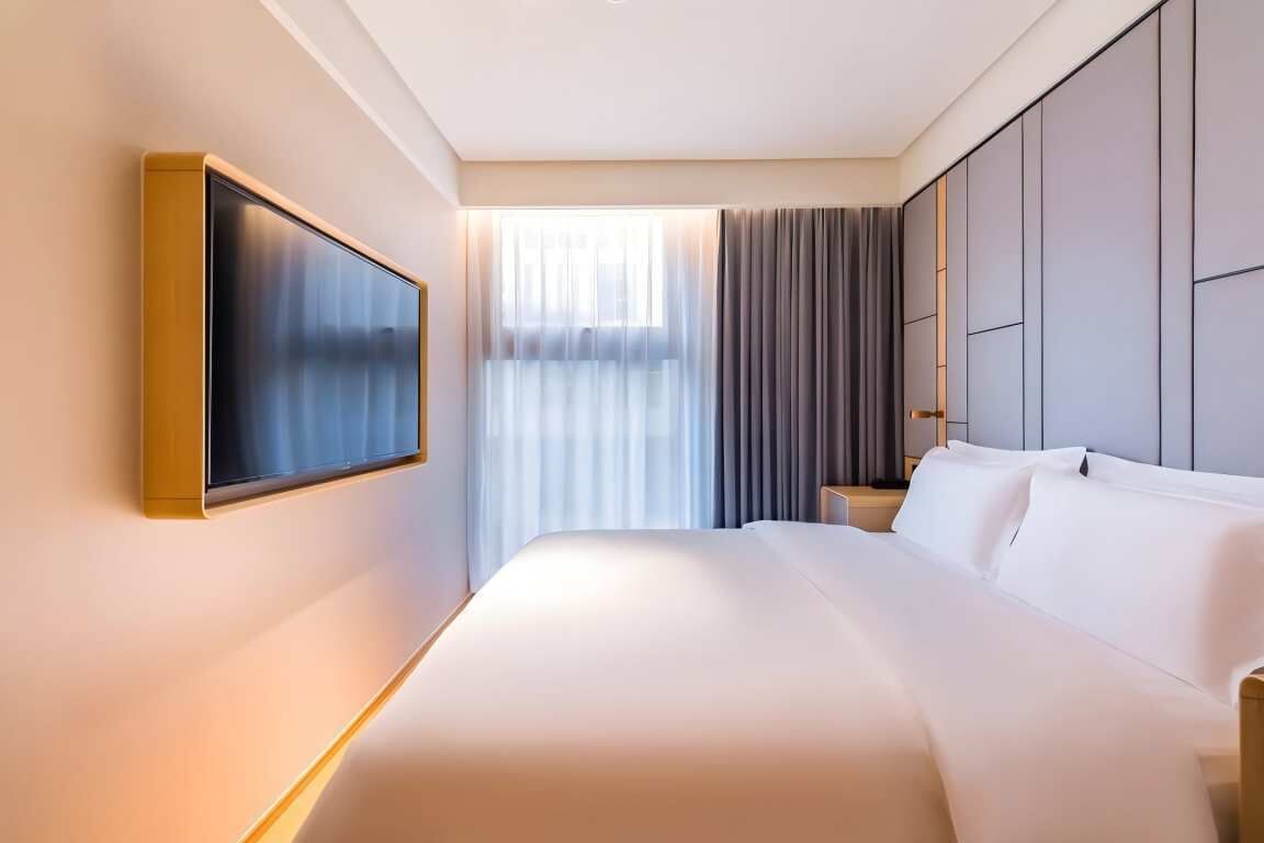 Quarto de hotel moderno com uma cama grande, uma televisão de tela plana na parede e uma janela ampla com cortinas.