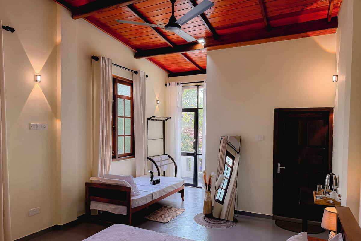 Quarto com cama, mesa de cabeceira, espelho, janela com cortinas, ventilador de teto e porta.