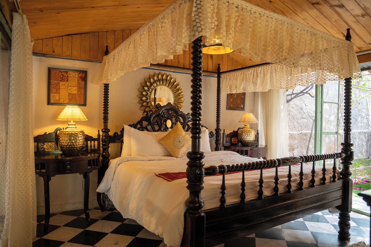 Uma cama com dossel e cortina, chão quadriculado, espelho com design em forma de sol e abajur em uma mesa de cabeceira.