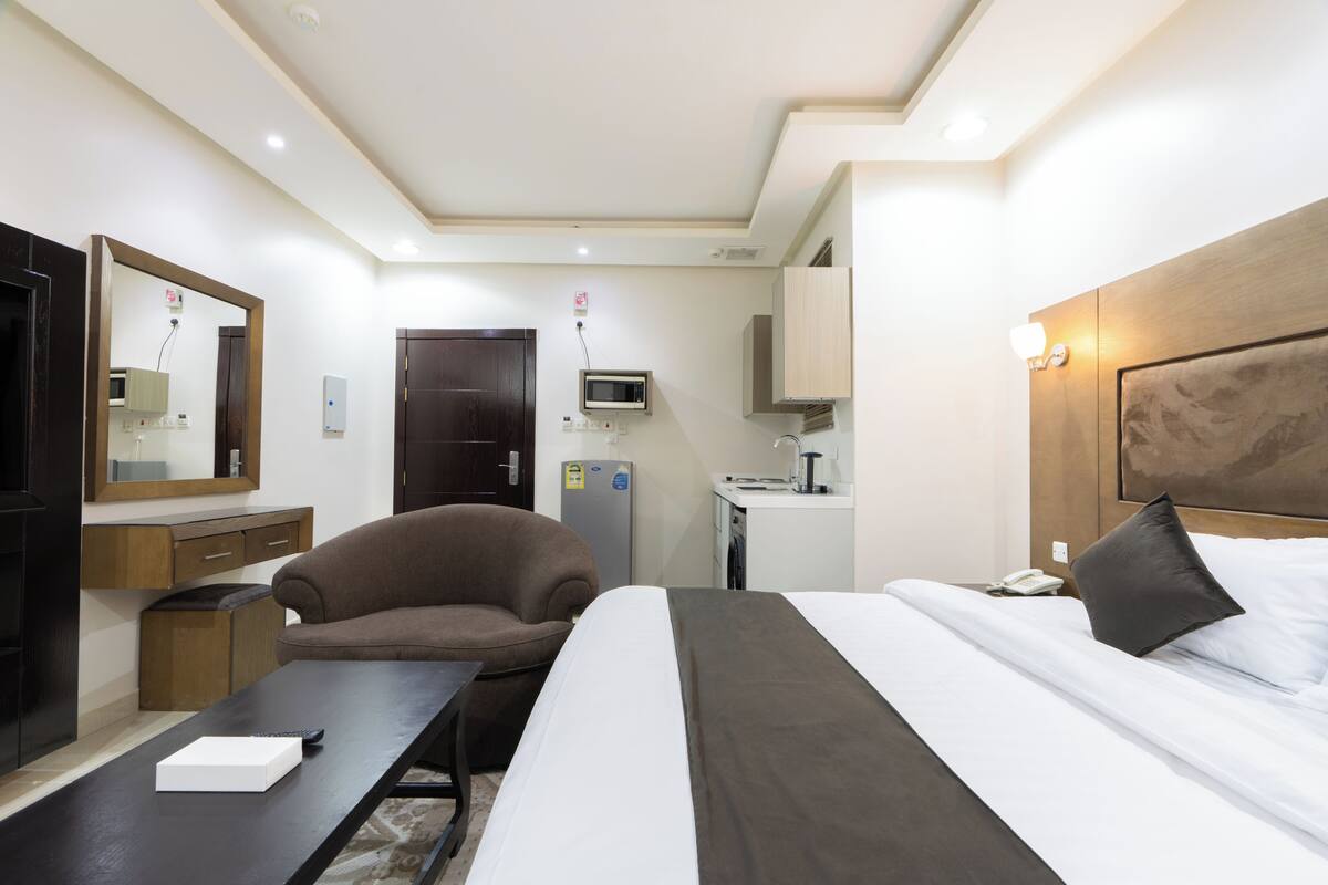 Quarto de hotel com cama, escrivaninha, cadeira, micro-ondas, frigorífico, espelho e mesa de cabeceira com telefone.