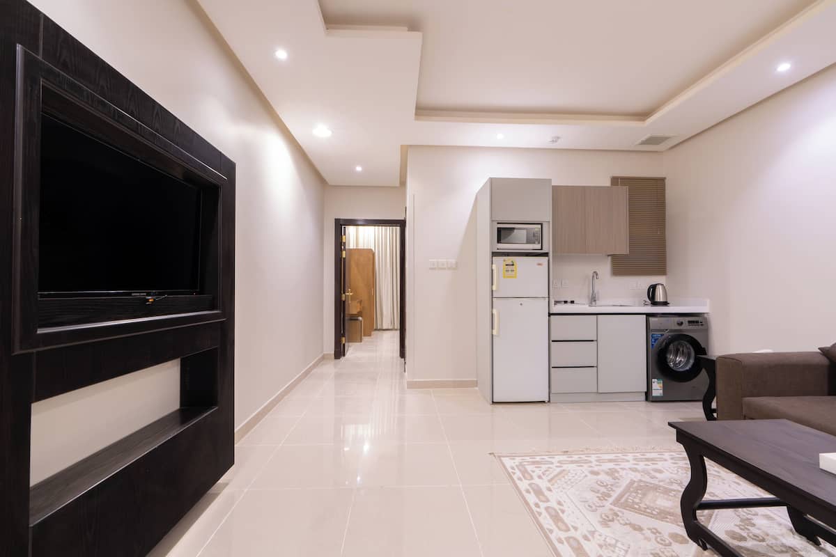 Uma sala de estar moderna com televisão de tela plana, sofá e mesa de centro. Uma área de cozinha com micro-ondas, forno e máquina de lavar roupa é visível ao fundo.