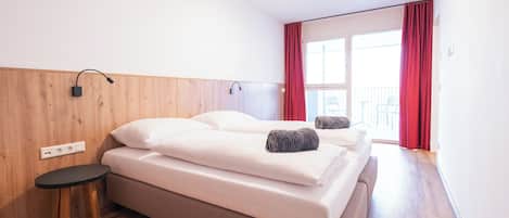 Quarto de hotel moderno com cabeceira de madeira, cama com lençóis brancos, um banquinho de madeira pequeno e uma janela com cortinas vermelhas.