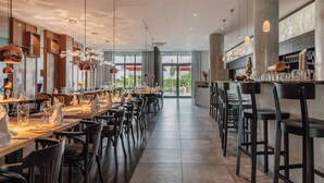Um restaurante moderno com mesas compridas, cadeiras pretas, luminárias pendentes e um bar com banquetas.