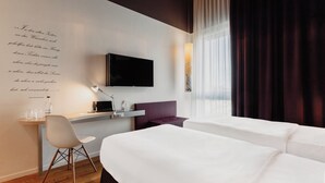 Quarto de hotel com cama, escrivaninha, cadeira, televisão e janela com cortinas.