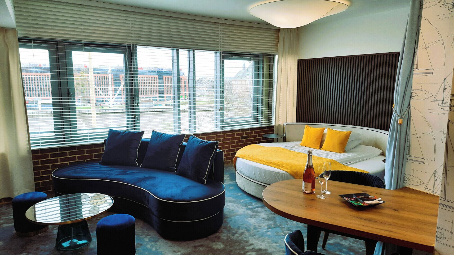 Quarto de hotel moderno com sofá curvo, cama com almofadas amarelas, mesa de madeira com uma garrafa e copos, e janelas amplas com persianas.
