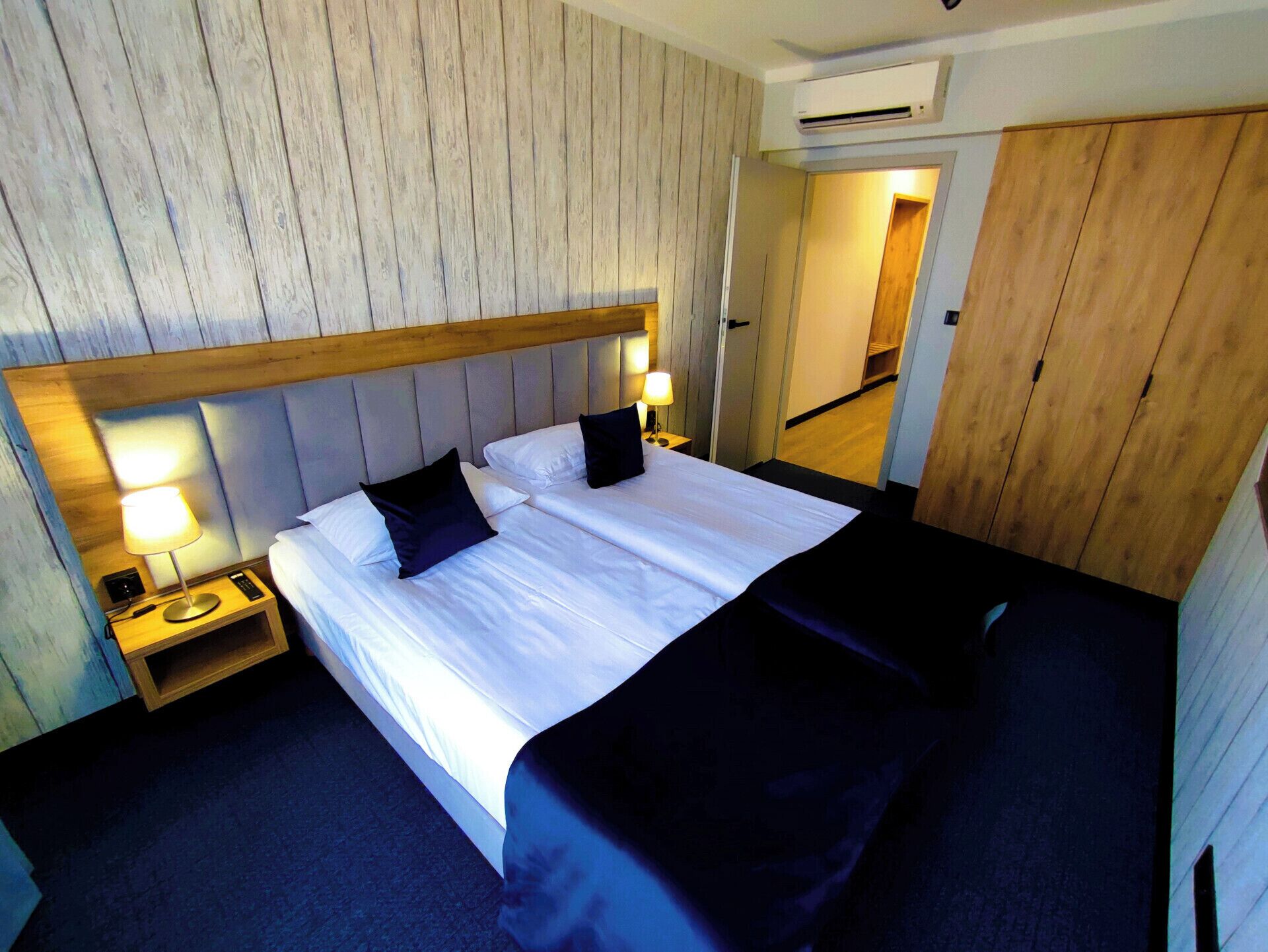 Quarto de hotel com uma cama grande, mesa de cabeceira com abajur, cabeceira de madeira e guarda-roupa.