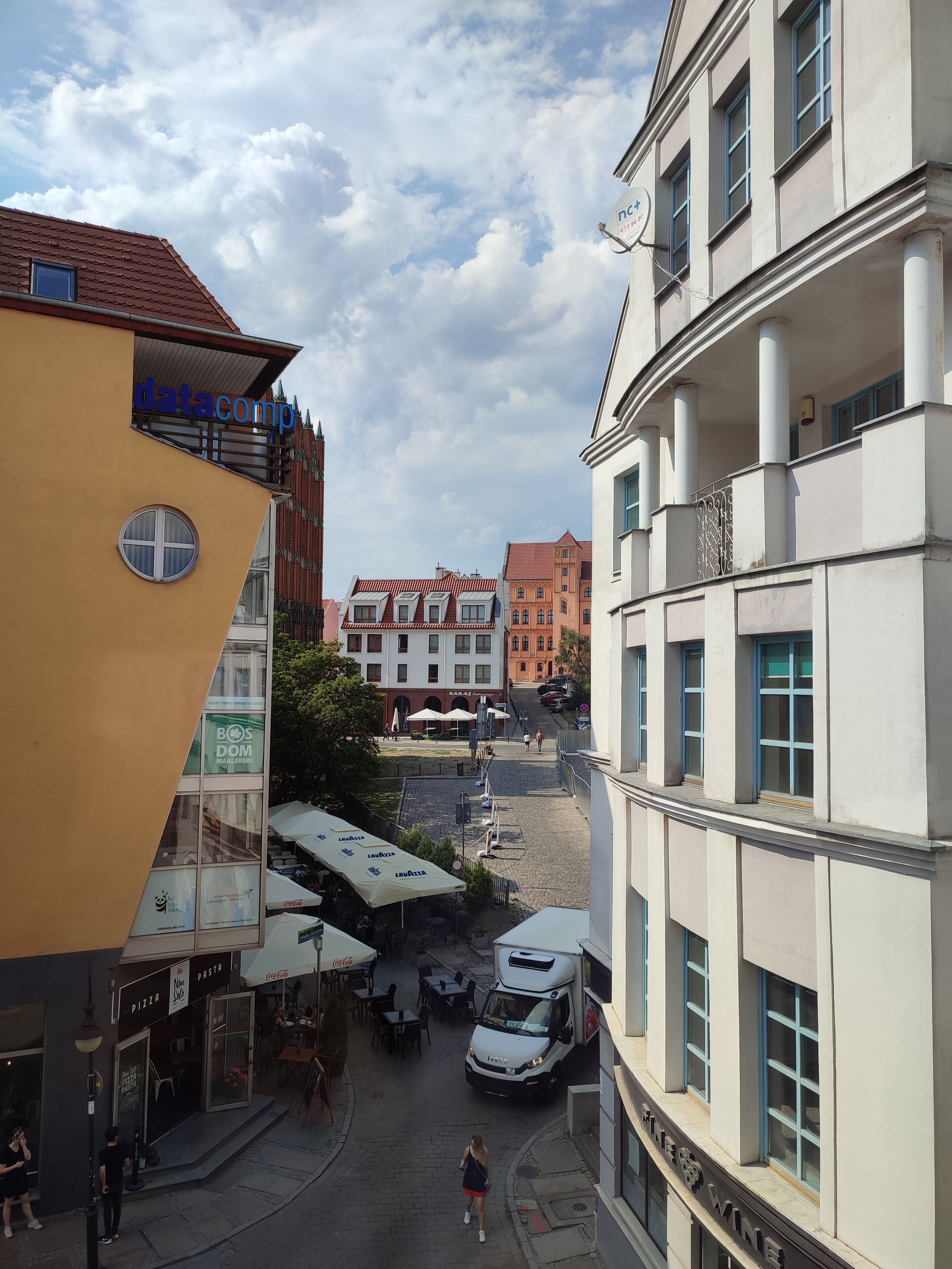 Vista da rua com edifícios, mesas ao ar livre e um veículo estacionado.