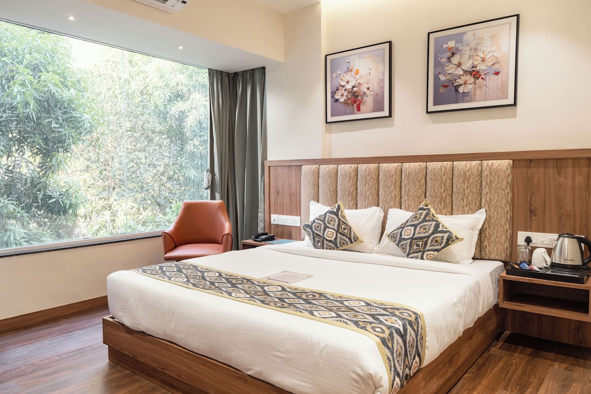 Quarto de hotel com uma grande janela, cama com roupa de cama estampada, cabeceira de madeira, mesa de cabeceira com chaleira e duas pinturas florais emolduradas na parede.