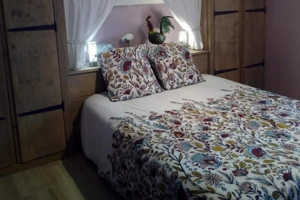 Quarto com cama, roupa de cama estampada com flores, guarda-roupa de madeira e janela com decoração de galo.