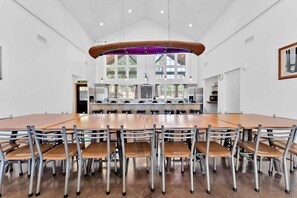 Uma sala de jantar ampla com uma longa mesa de madeira e cadeiras de metal, janelas grandes e teto alto com iluminação pendente.