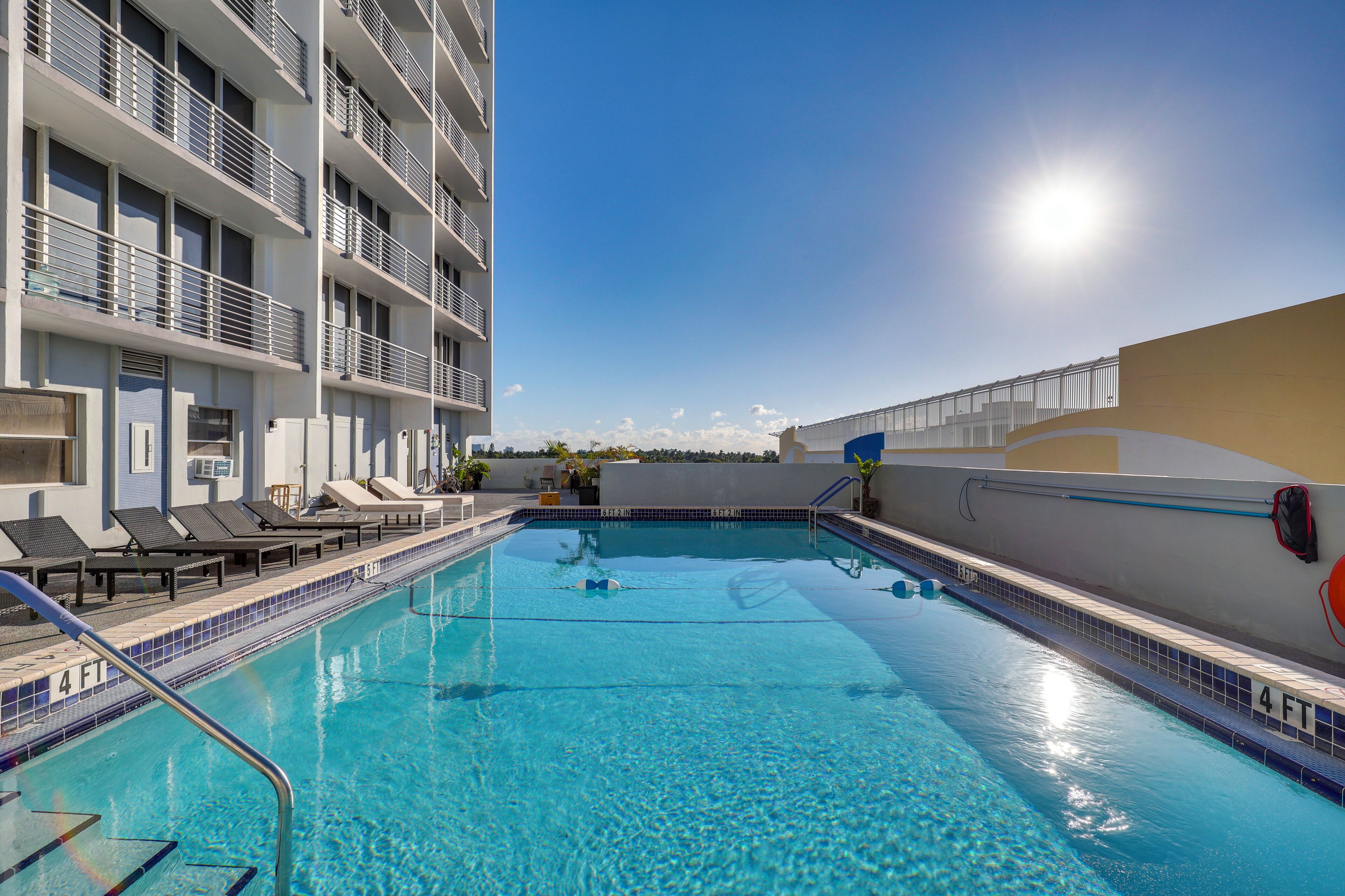 Uma piscina sob um céu ensolarado, um edifício com varandas e espreguiçadeiras.