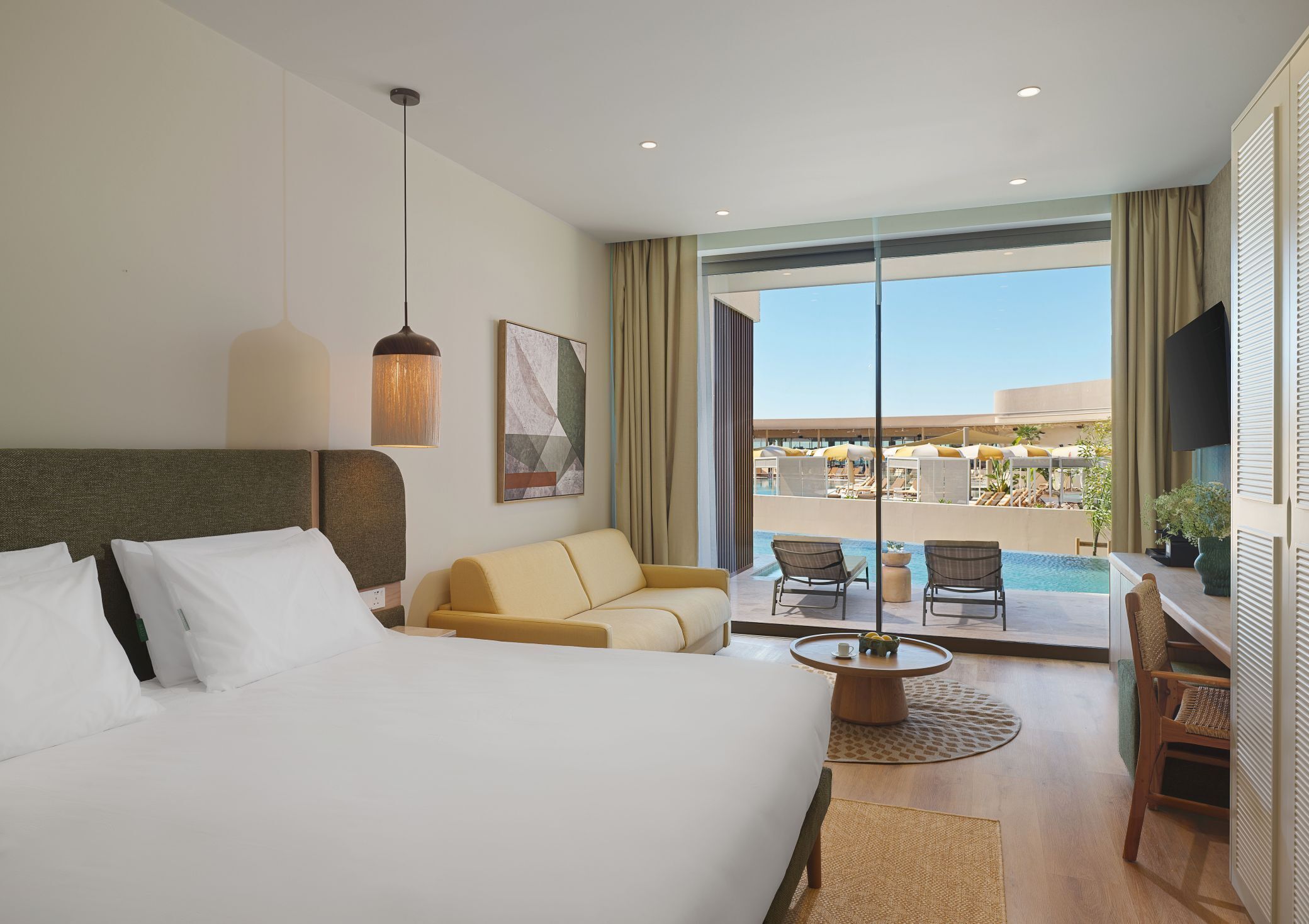 Quarto de hotel com cama, sofá, mesa de jantar, TV e vista para piscina e área externa.
