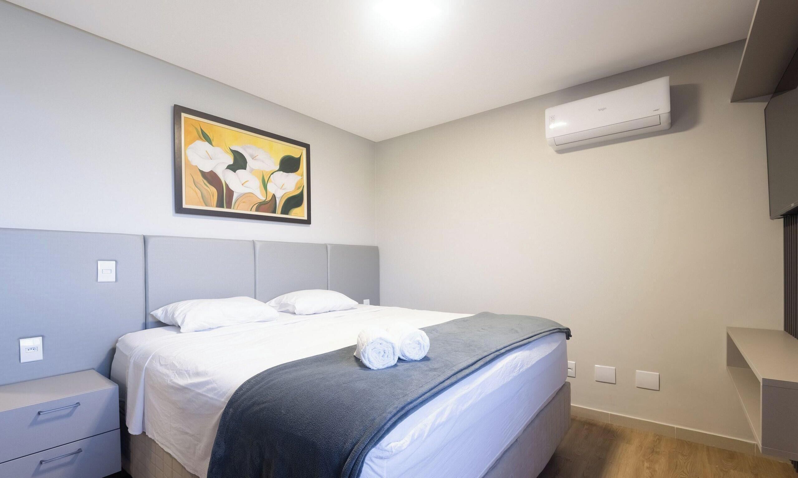 Quarto de hotel moderno com cama, mesa de cabeceira, maleiro, quadro e aparelho de ar condicionado.