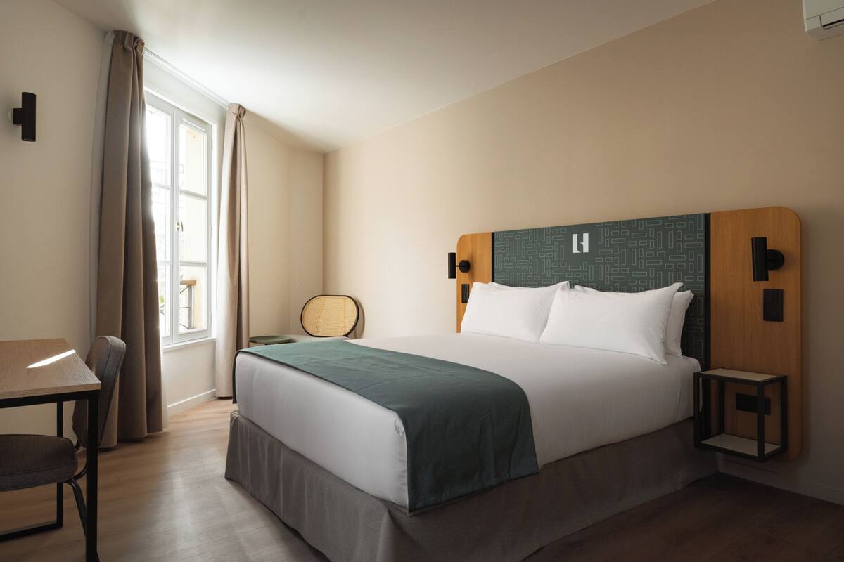 Quarto de hotel com cama, uma cadeira, uma mesa e janela com cortinas.
