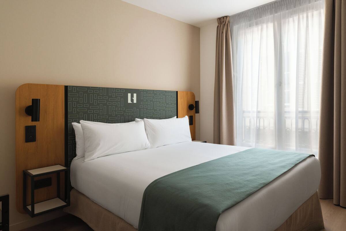 Quarto de hotel com uma cama, mesa de cabeceira, painel de controle fixado na parede e uma janela com cortinas.
