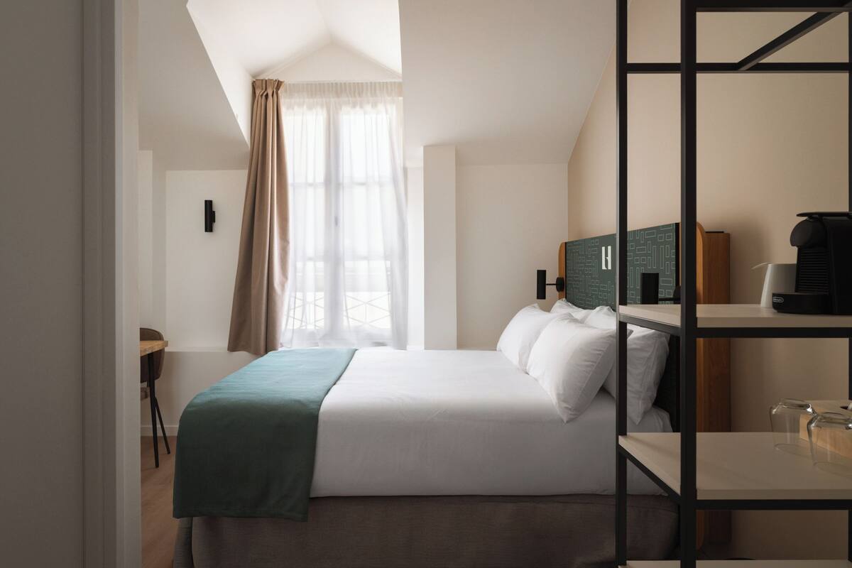 Quarto com cama, escrivaninha, cadeira, máquina de café e janela com cortinas.